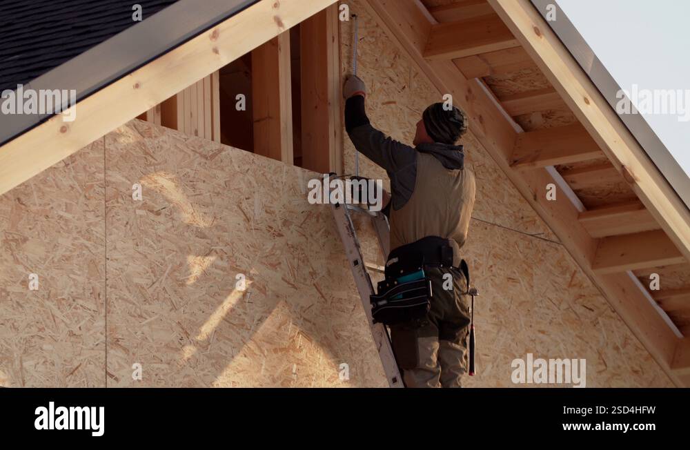 Worker is uses measuring tape standing on ladder. Frame house under ...