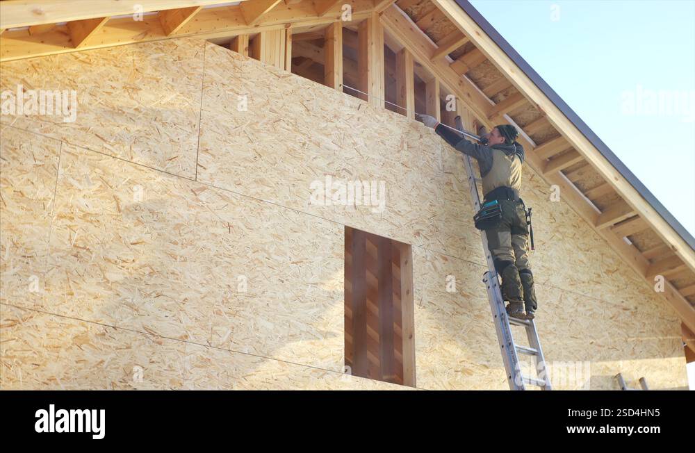 Worker is uses measuring tape standing on ladder. Frame house under ...