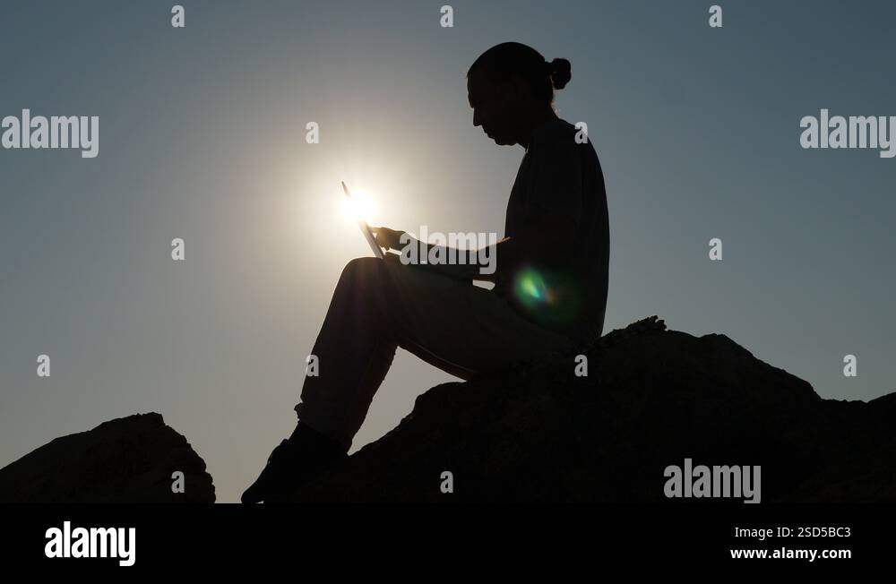 Shadow figure of guy sit on stone. He using laptop, swipe touch screen ...