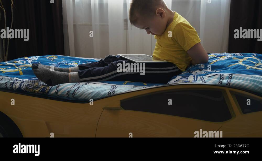 Boy sits on bed, tablet lies on his lap, hunched over looking at screen ...