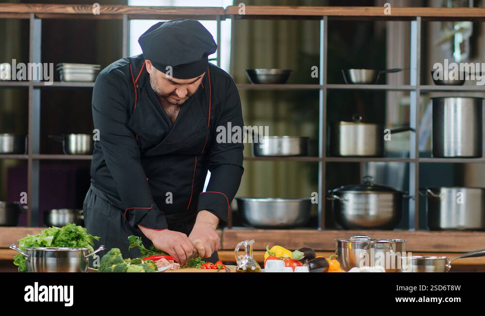 Handsome male chef in black uniform cutting greens and pepper. Medium ...