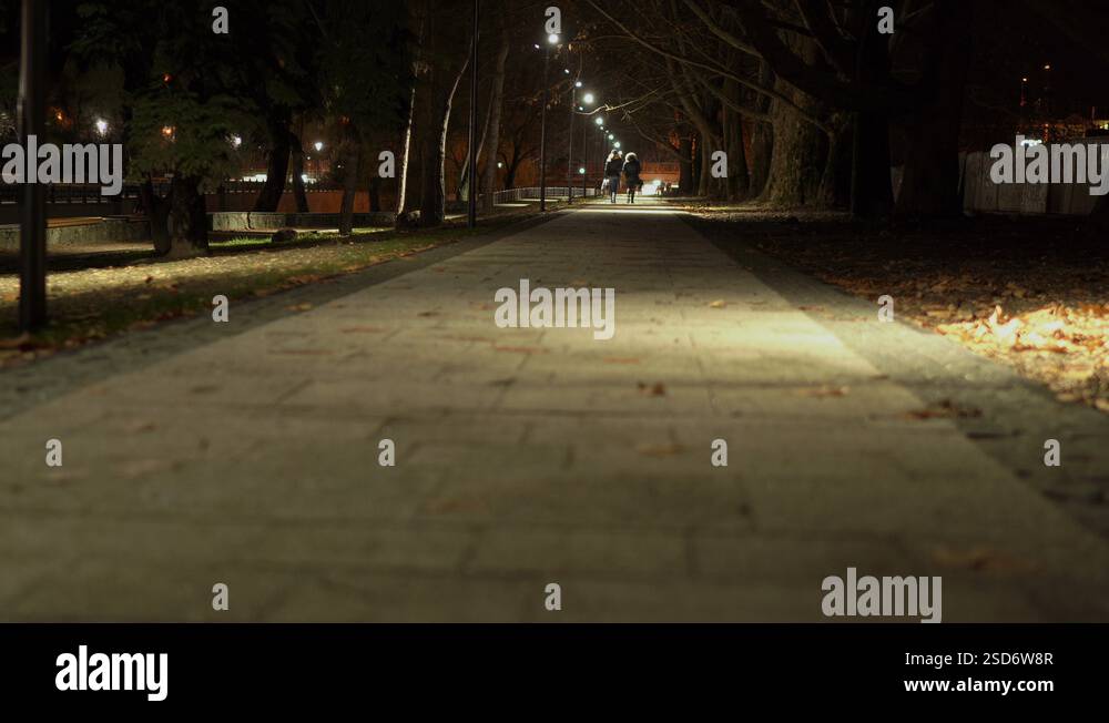 Two women walking down a paved footpath lit by lanterns at night in ...
