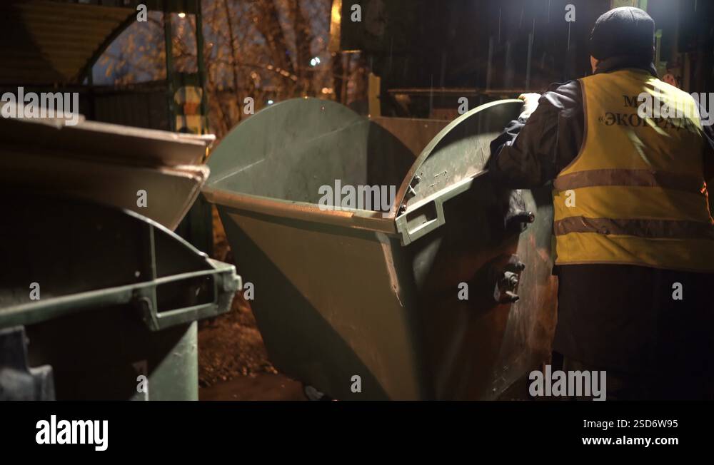 Garbage man at work on rainy city street at night, garbage truck lifts ...