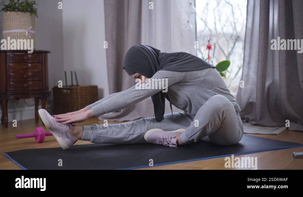 Side view of slim Muslim woman bending forward on yoga mat trying to ...