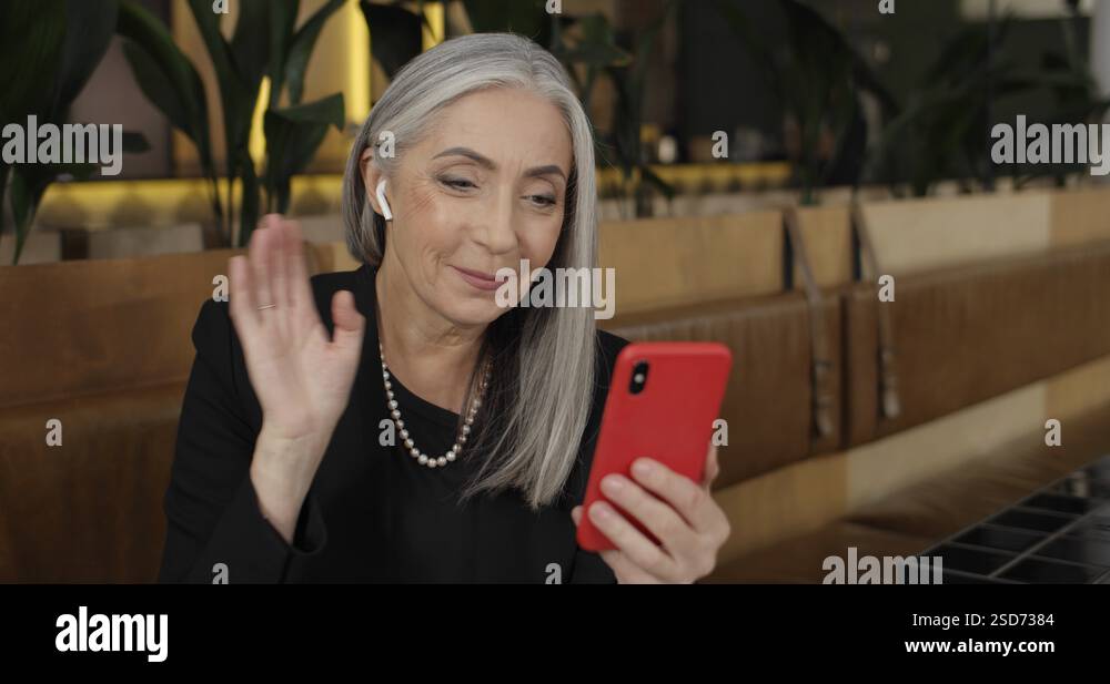 Older good looking woman in earpods waving to smartphone camera ...
