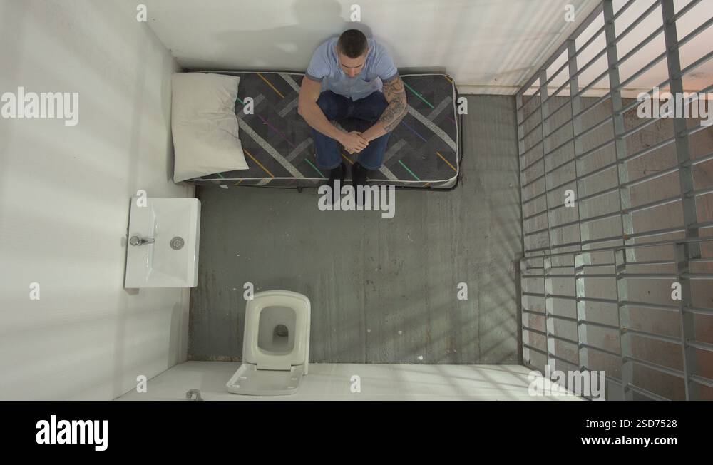 Male Prisoner in Prison Cell. The inmate is sat on his bed. Overhead 4K ...