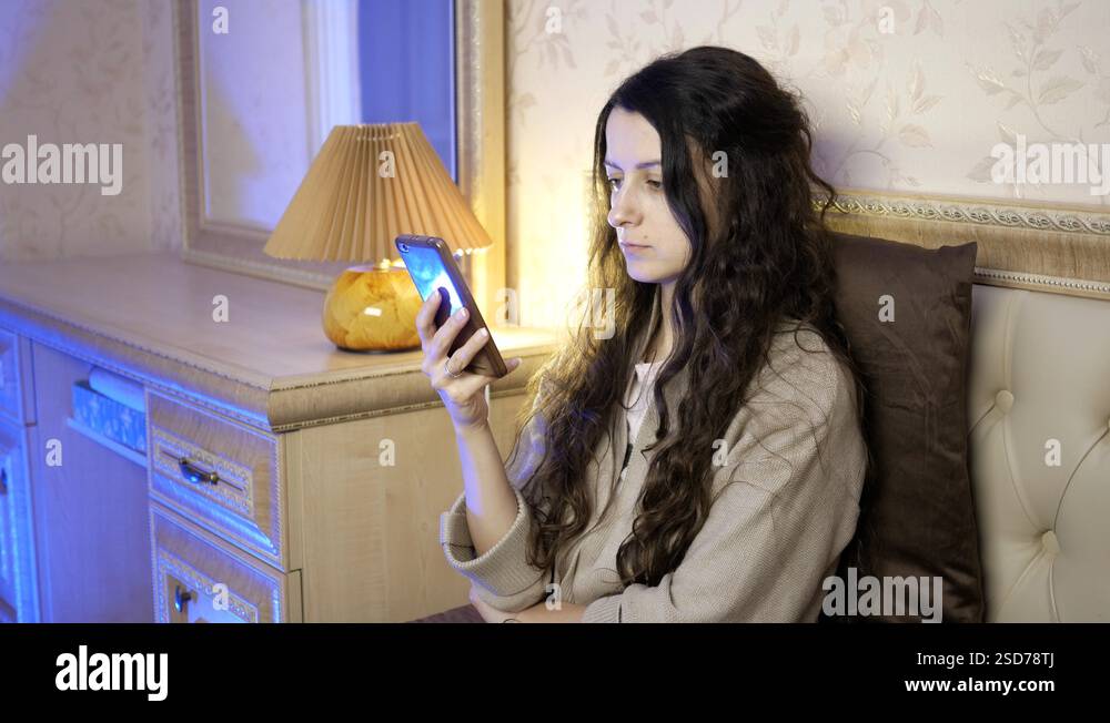 Beautiful young woman uses mobile phone while sitting on bed in bedroom ...