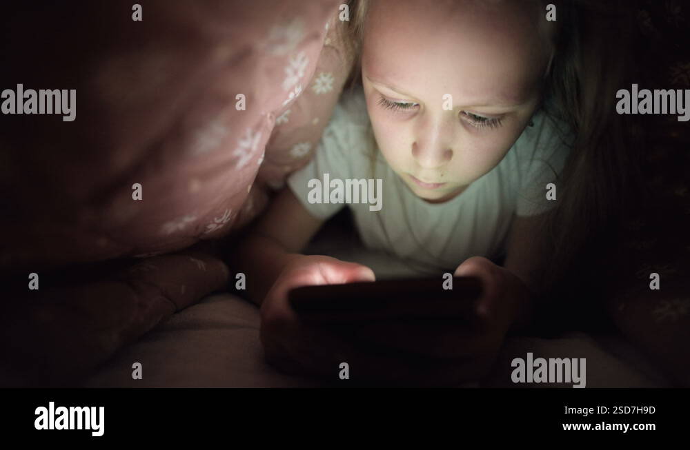 A young little girl is hiding under a blanket to use a digital tablet ...