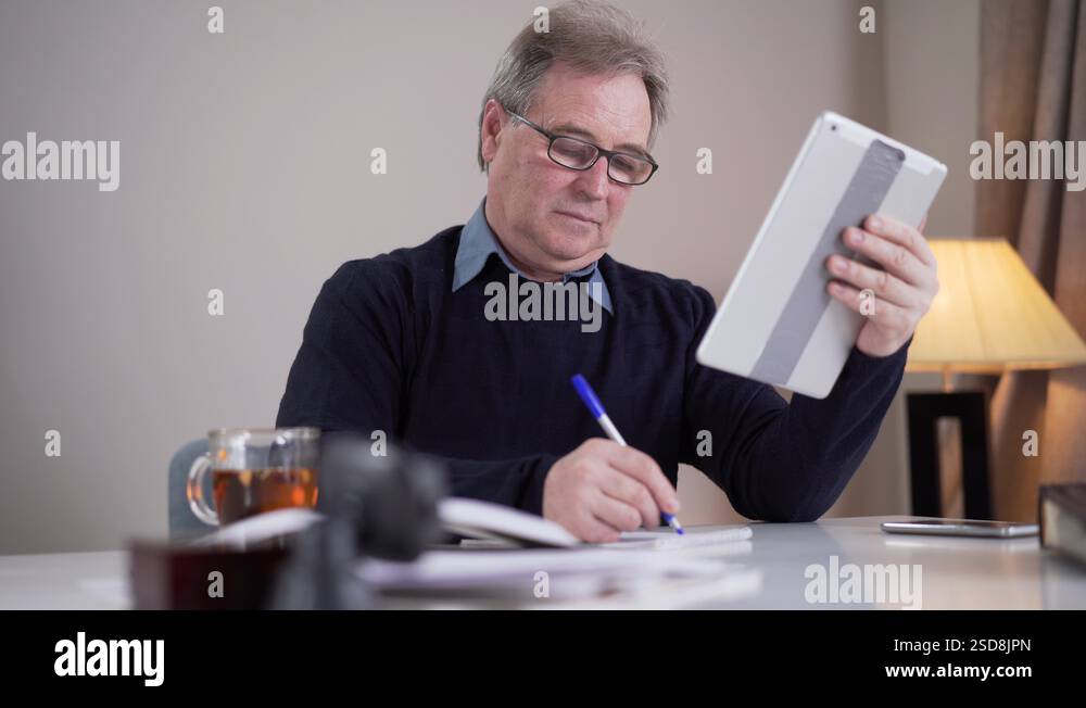 Senior Caucasian man writing at the table when having incoming video ...