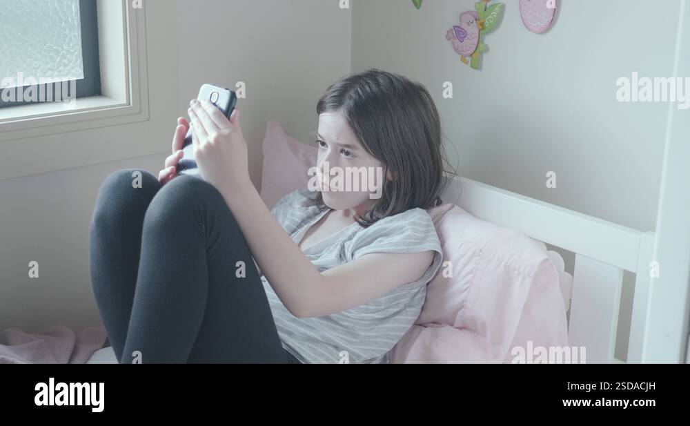 young girl concentrates intently on her mobile phone while on her bed ...