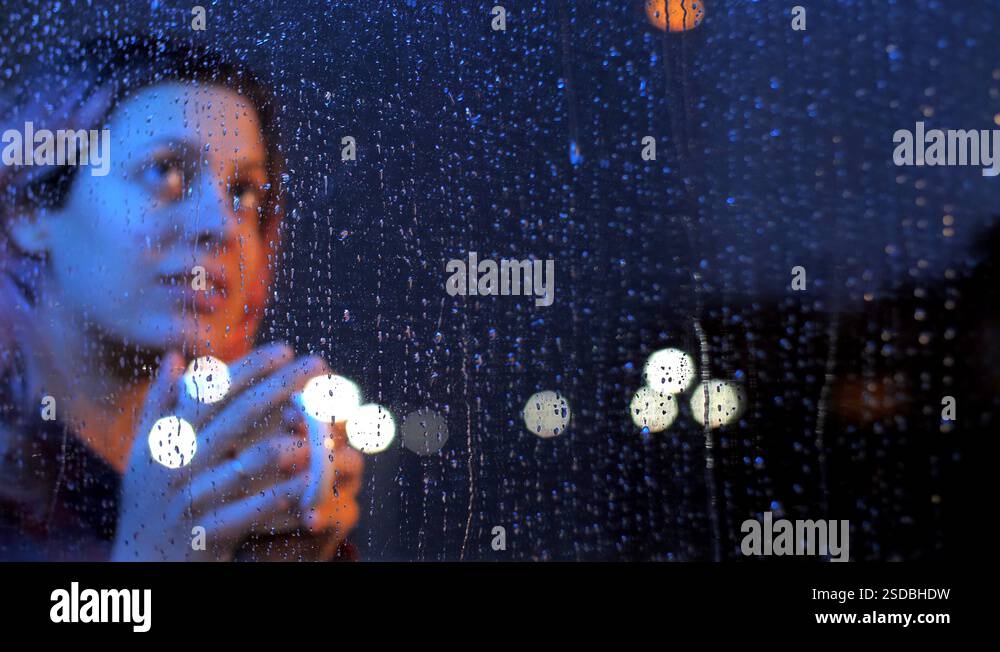 Woman drinks hot tea or coffee from a mug and looks out on raining ...