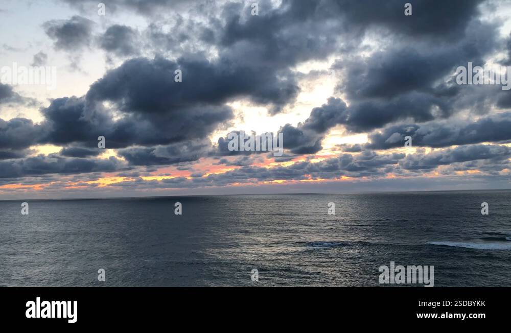 Gray and blue clouds move over the ocean at sunset with oranges and ...