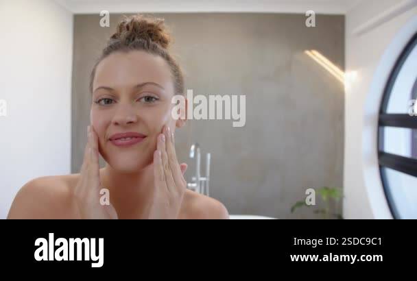 Applying skincare, young woman smiling and touching face in bathroom ...