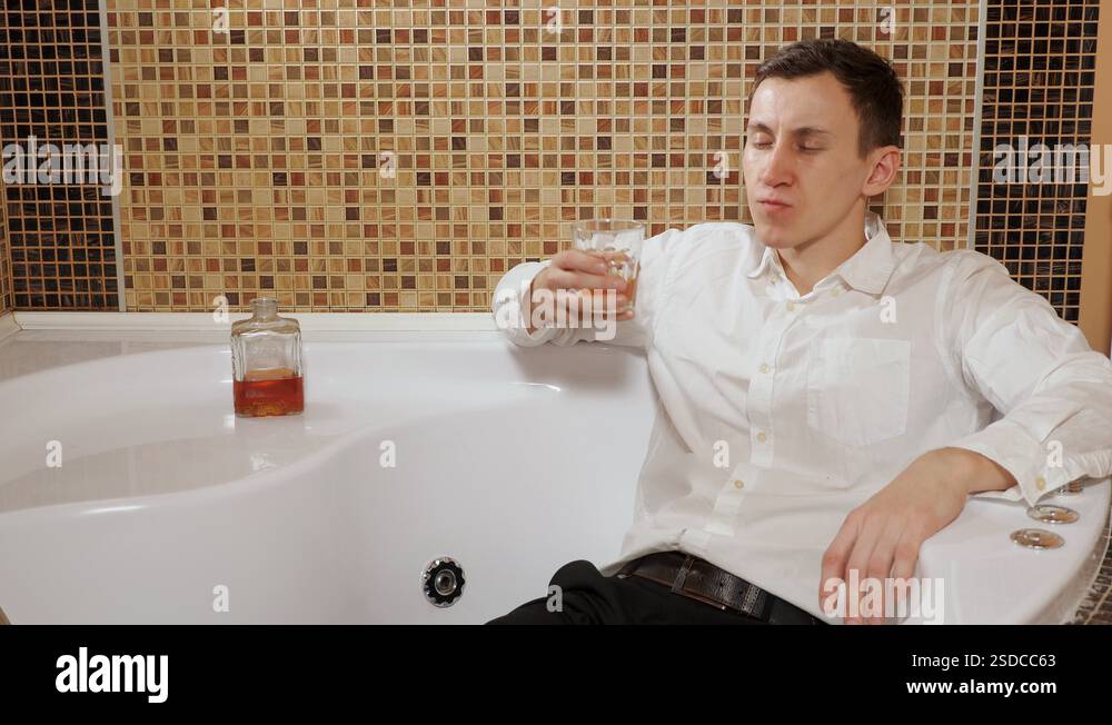drunk man in trousers and a shirt is lying in the bath and drinking ...
