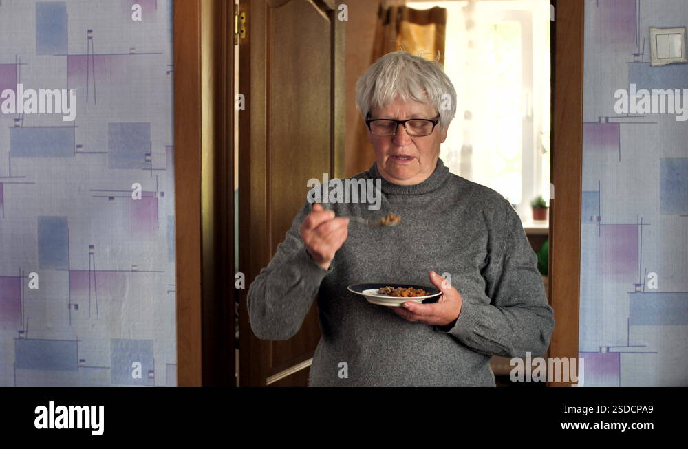 gray-haired old woman is eating poor food with a spoon from a plate ...