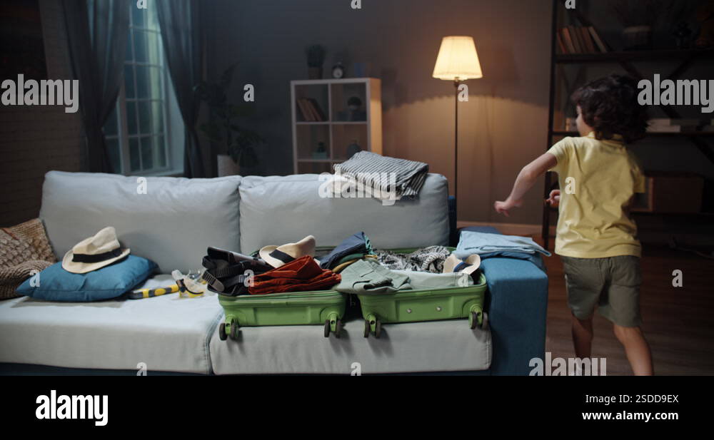 Funny little asian kid with curly hair is hurryingly packing a suitcase ...