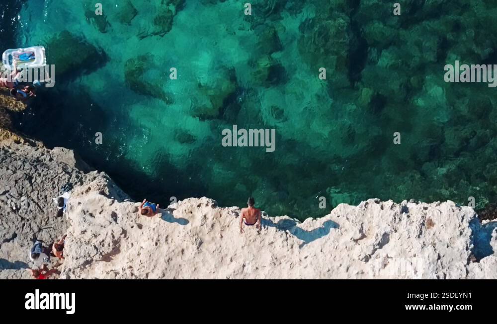 Slow motion aerial top down shot of man jumping from a cliff into clear ...