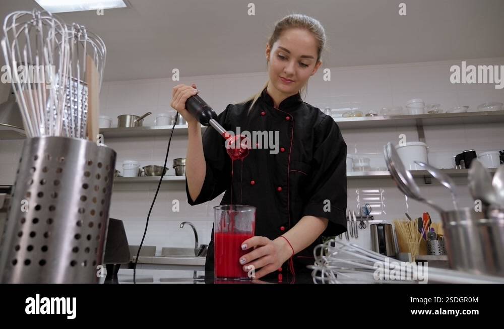 A female pastry chef prepares berry sauce or mousse using a blender in ...