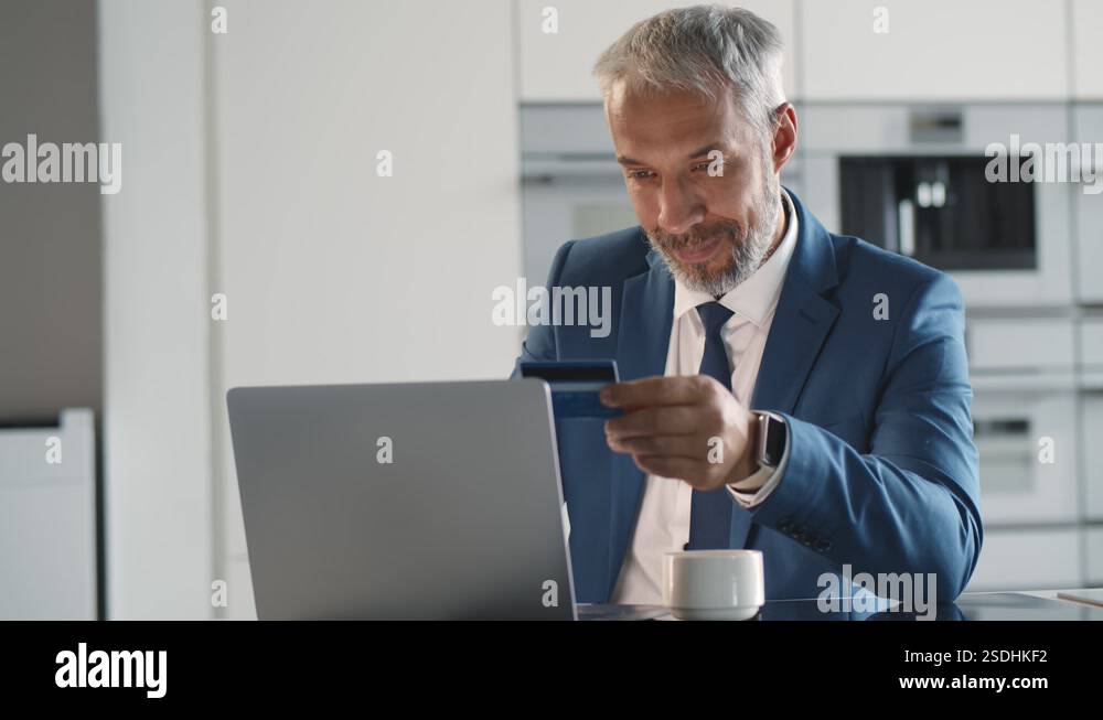 Senior businessman sitting in kitchen, purchasing on internet and ...