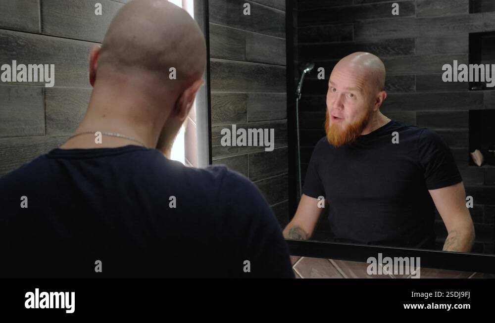 A young bald guy with a red beard points his finger at his reflection ...