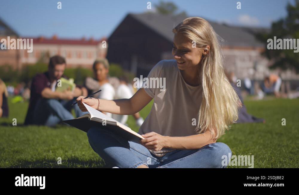 Beautiful young female student reading book outdoors sitting on lawn i ...