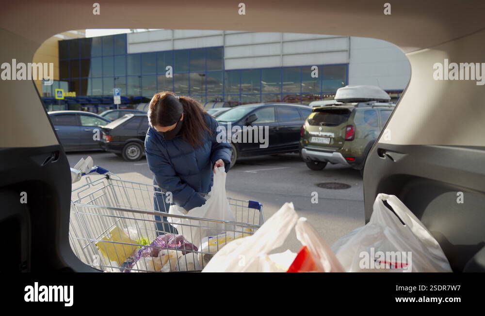 Woman in protective mask near car loads groceries in trunk on shop ...