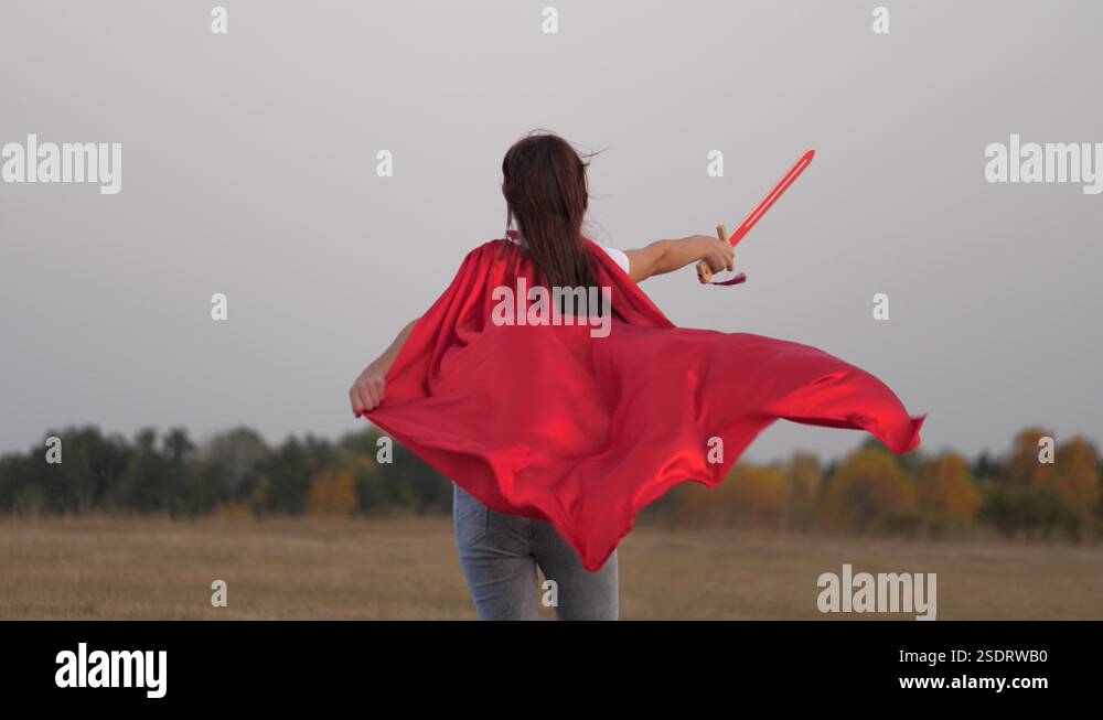 Cheerful girl in red cloak runs with swords in his hand across field ...