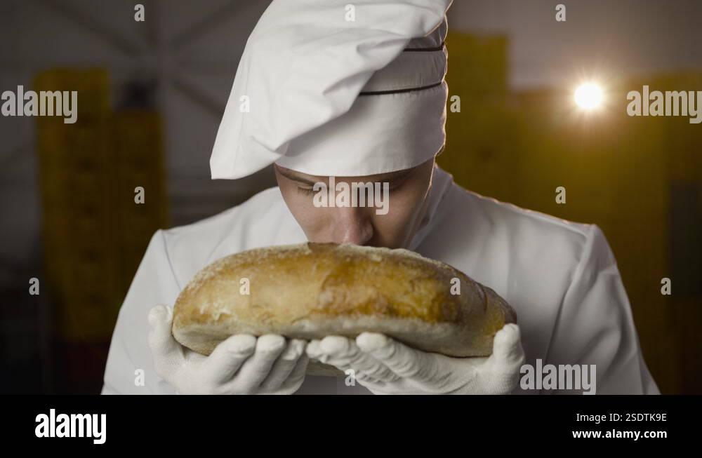 Baker in uniform sniffs a loaf of bread, has a pleasure and smile at ...