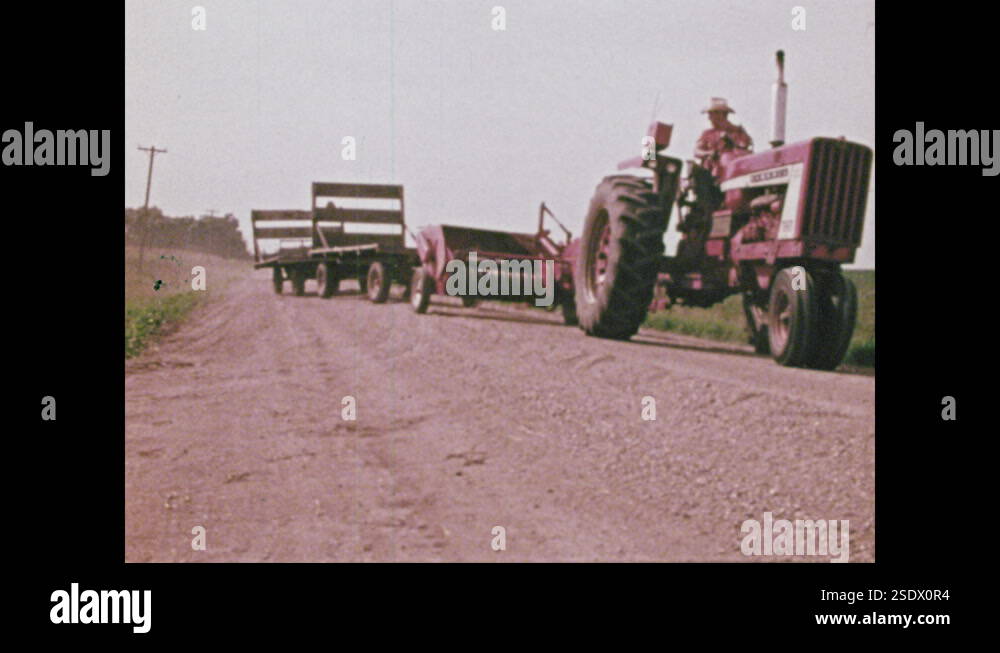 1970s: Man drives tractor pulling trailer on farm. Man adjusts trailer ...