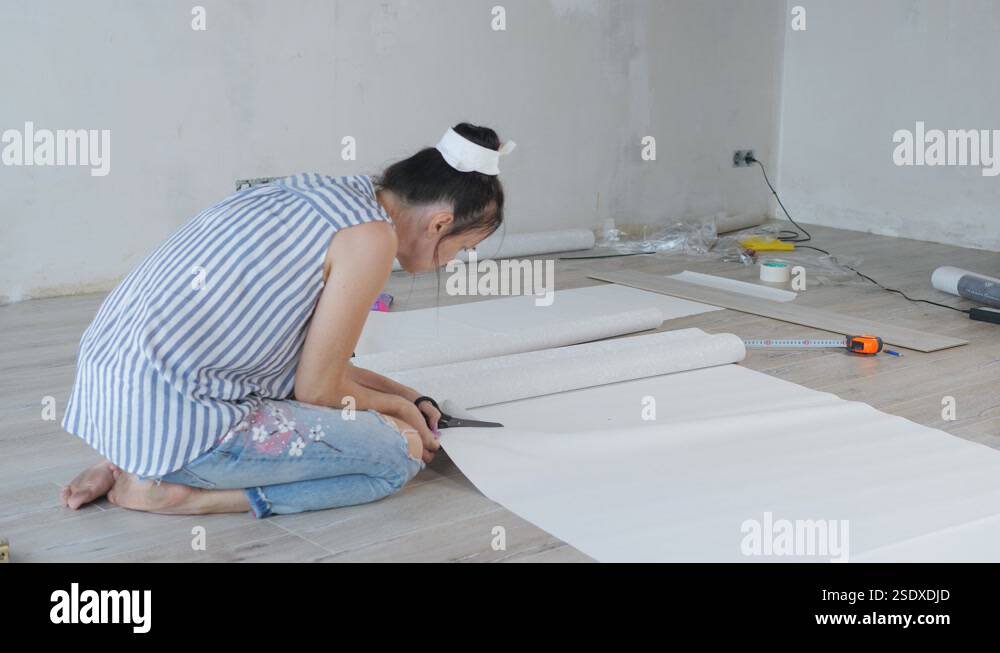 A woman on the floor cuts off a sheet of Wallpaper with scissors ...