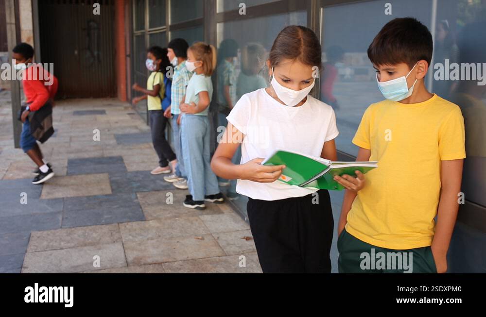 Boy and girl in face masks looking at exercise book talking about ...