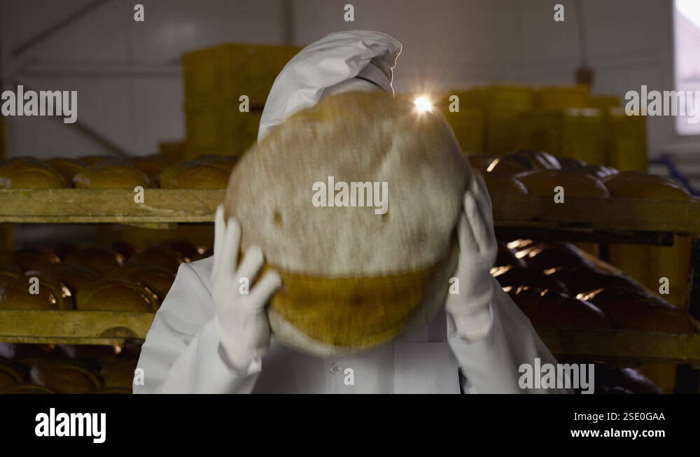 Young baker in uniform raises large loaf of round bread at face level ...