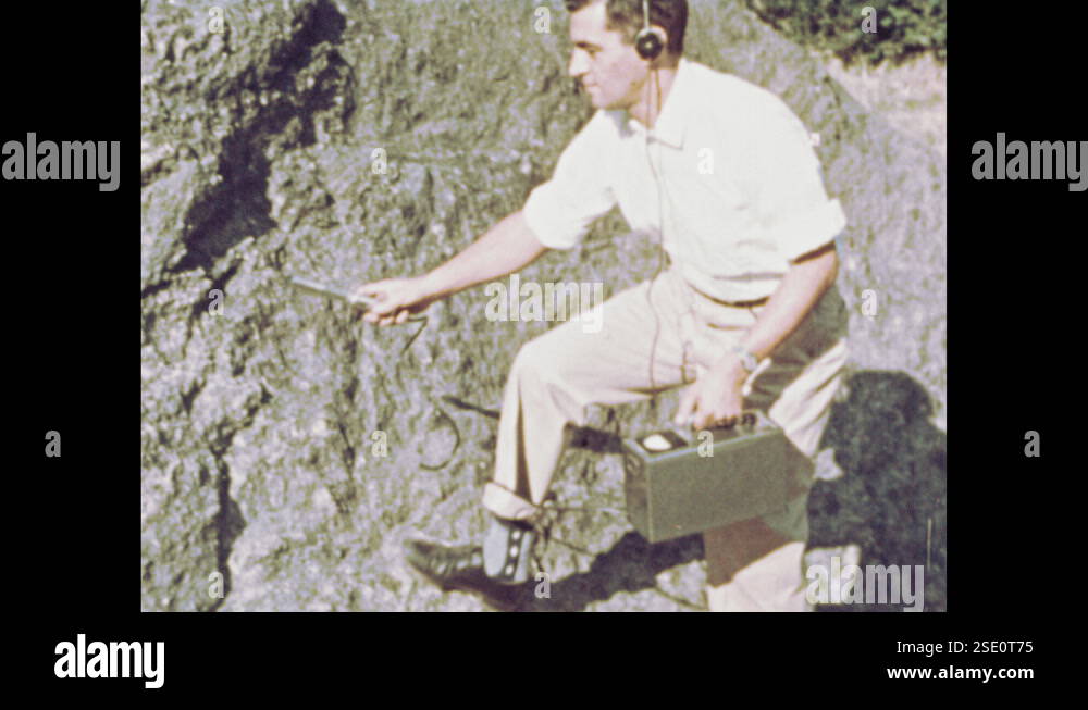 1960s: Man passes Geiger counter over rocky surface. Exterior atomic ...