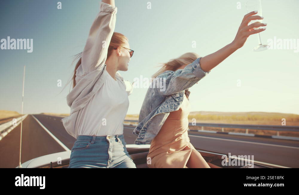 Two beautiful young women fun ride in convertible raising their hands ...