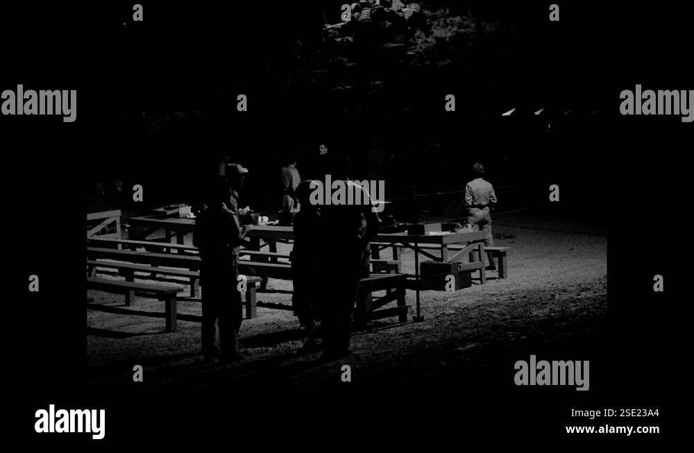1940s: Soldiers stand around outside benches and tables at night. Two ...