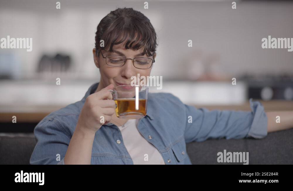 Young woman wearing glasses sitting alone at her home drinking herbal ...