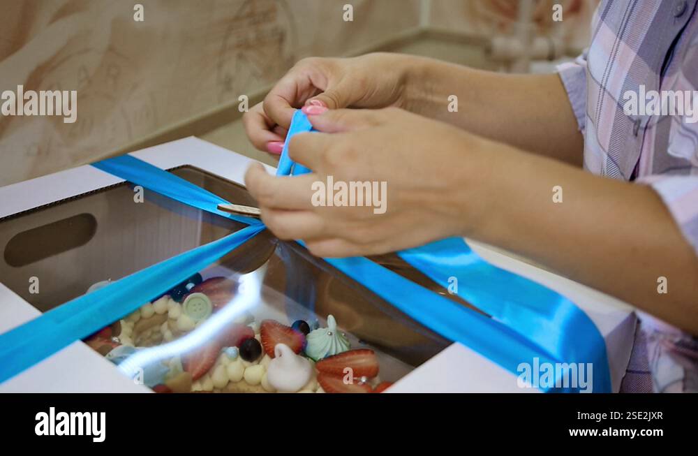 Young woman pastry chef in the kitchen tying blue ribbon on white cake ...