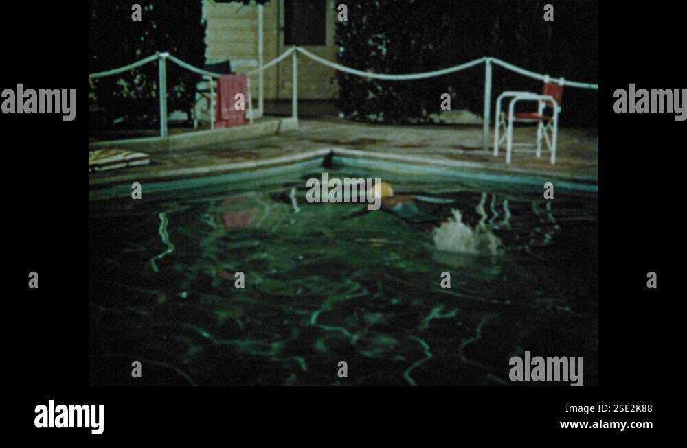 1950s: Man on diving board over pool reaches hand to man in pool. Woman ...