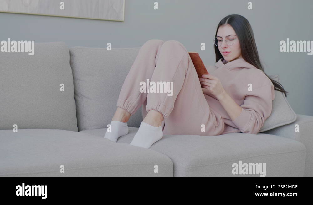 A young Caucasian woman wearing reading glasses reading book on sofa ...