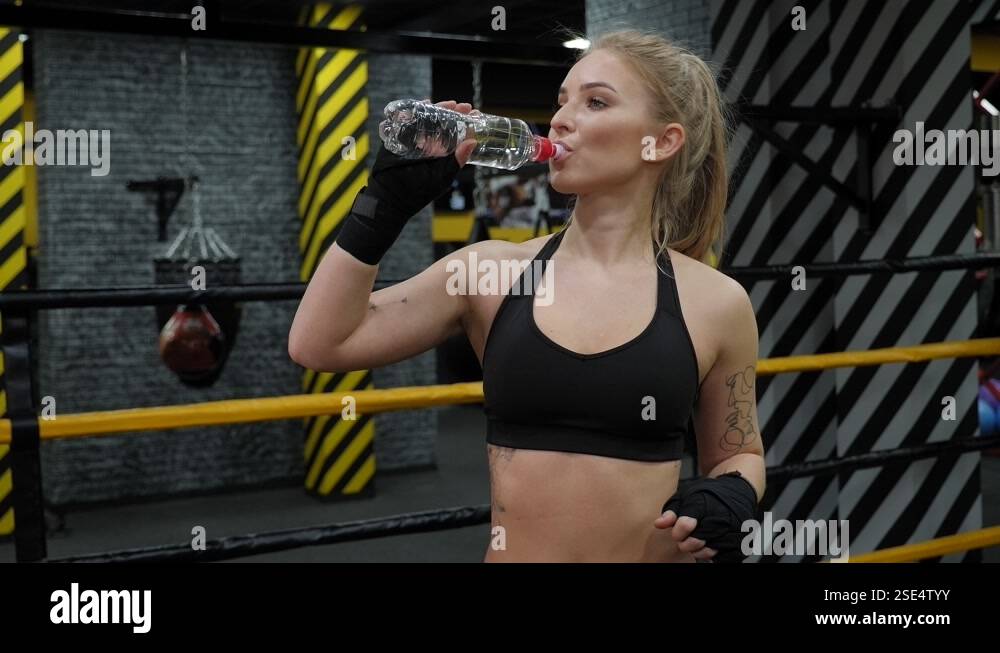 A tired girl drinks water standing in the boxing ring. Boxing training ...