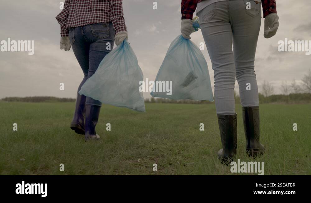 Girls walk on a green field in rubber boots and hold garbage bags in ...
