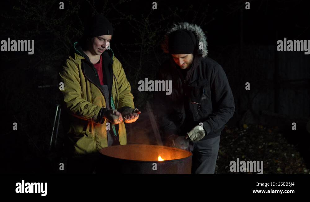 Two homeless young men bask in the fire. Men stand near a barrel of ...