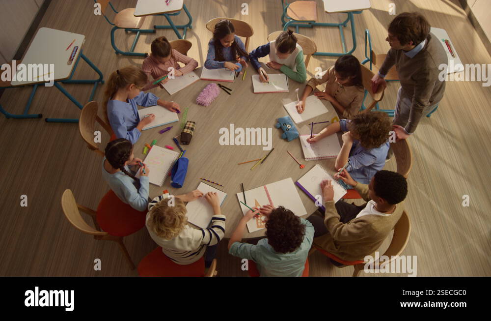 Schoolchildren having lesson at round table. Male teacher helping kids ...