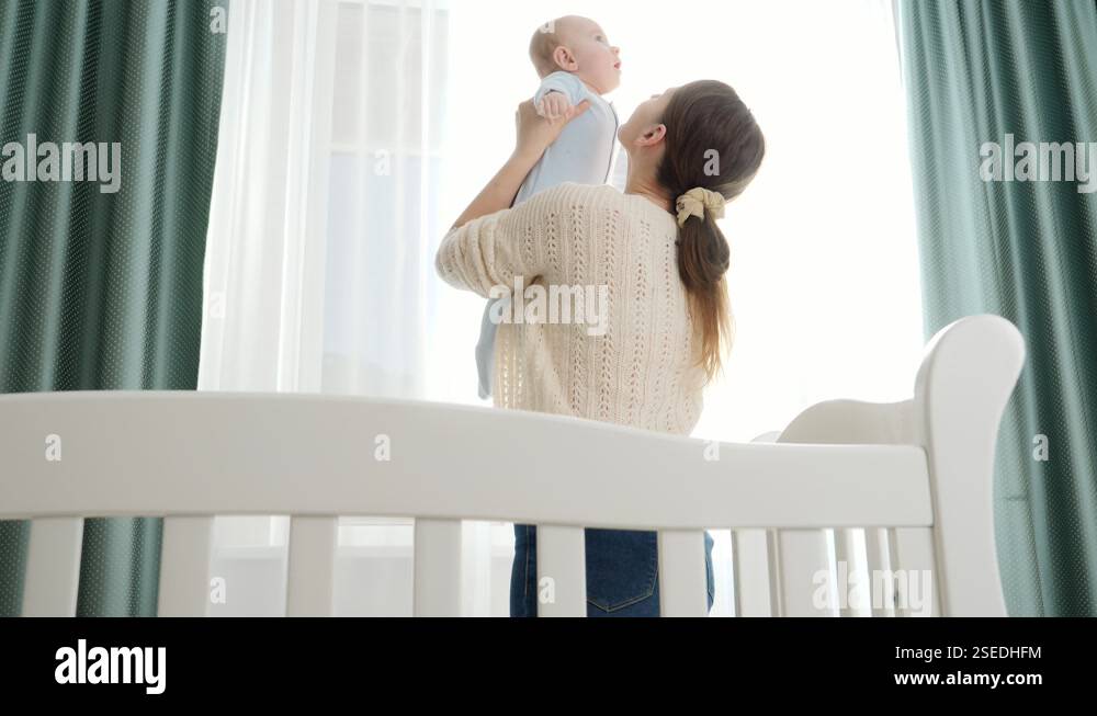 Young mother standing at cradle and holding her smiling baby son ...