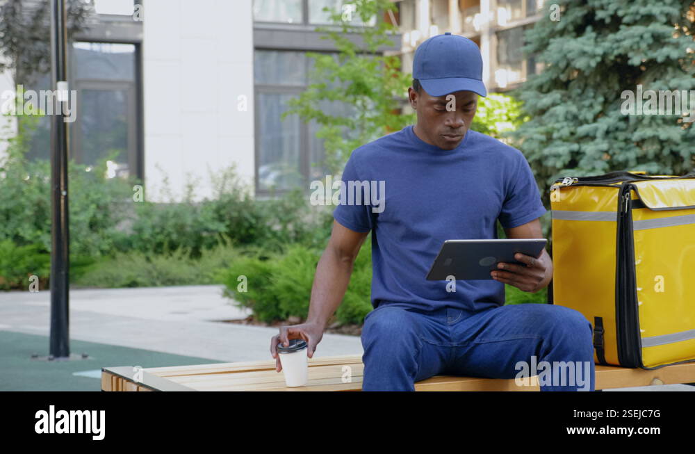 African man courier food delivery sitting on bench uses tablet drinking ...