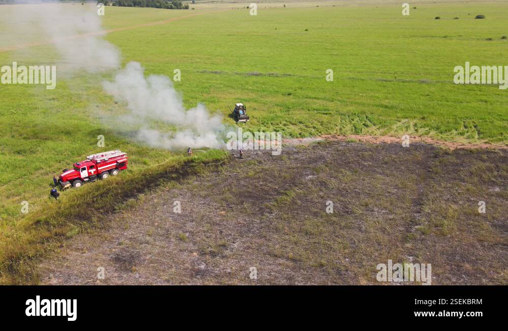 A fire truck and a tractor on the edge of a burnt field with smoke ...