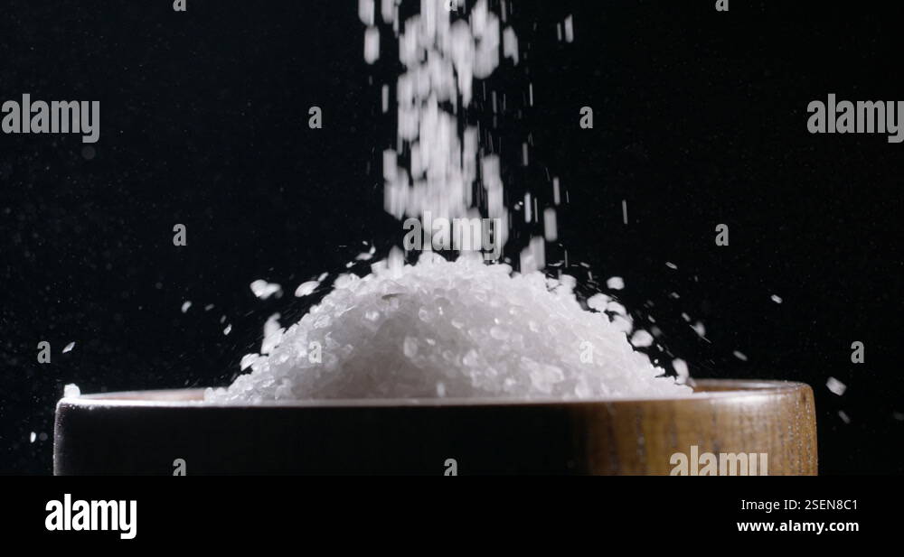 White table salt granules falling on wooden surface and forming a pile ...