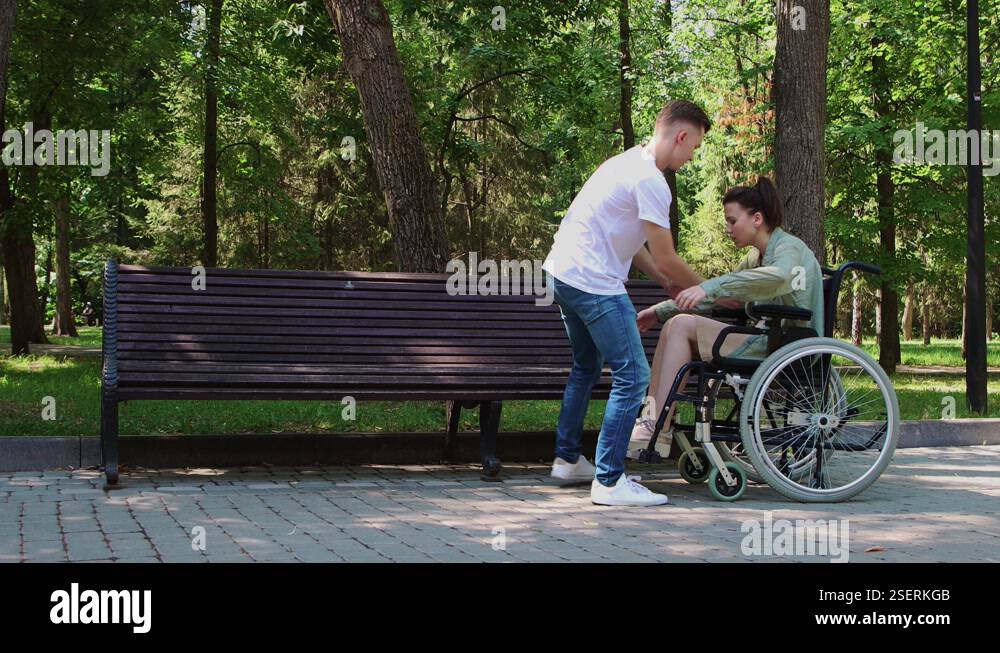 A guy helping disabled woman to sit from the wheelchair to the bench ...