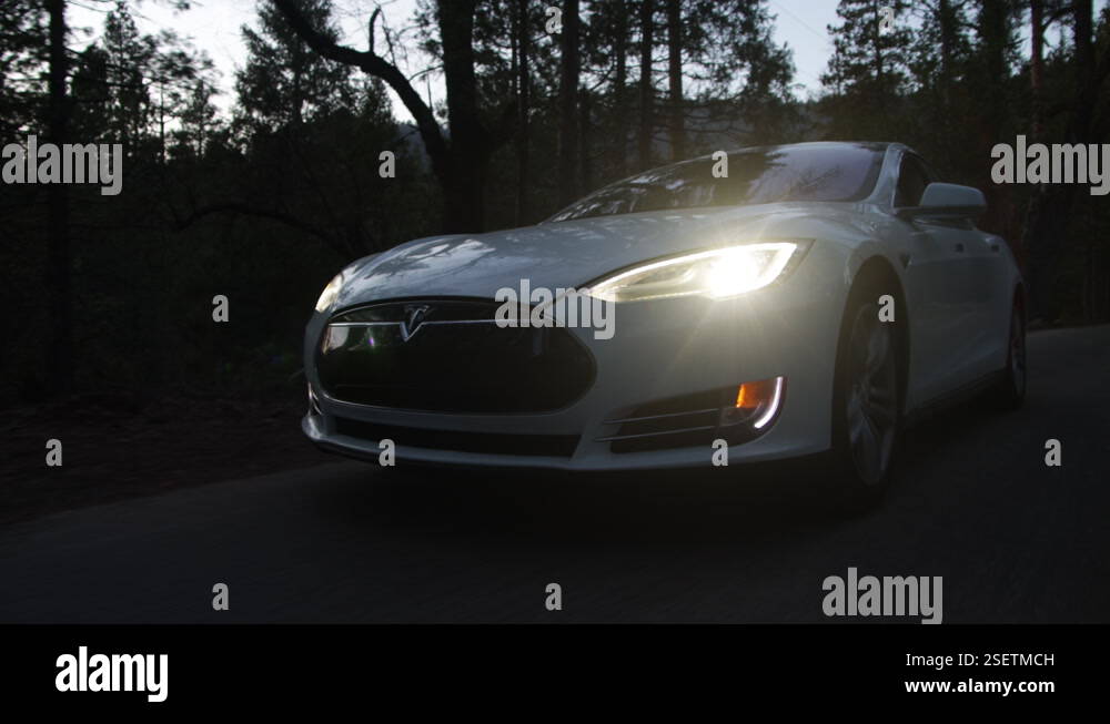 Close up front view of a Tesla car driving slowly in the dark on a road ...