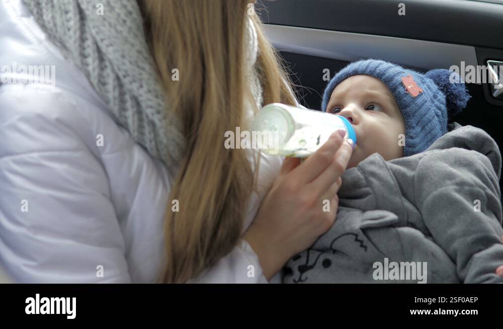 A mother feeds a one-year-old child in the back seat of a car with milk ...